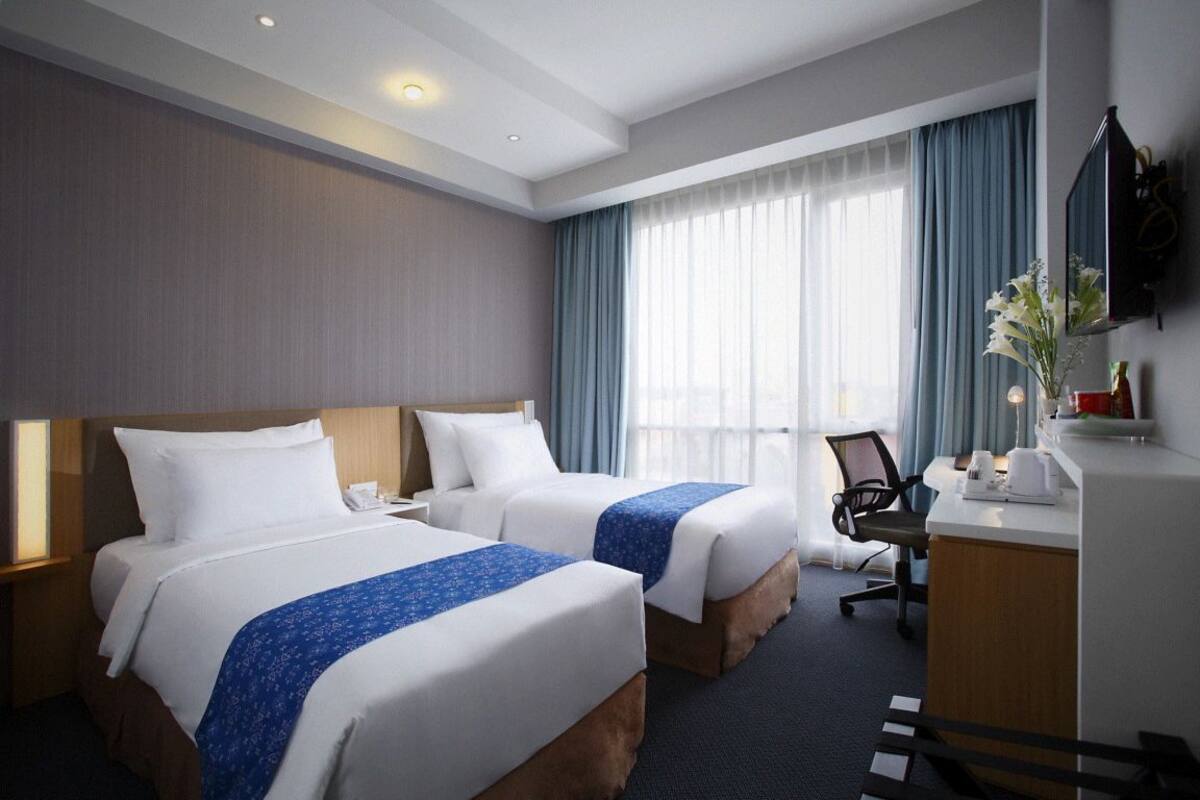 Quarto de hotel com duas camas, uma escrivaninha, uma cadeira, televisão e janela com cortinas.