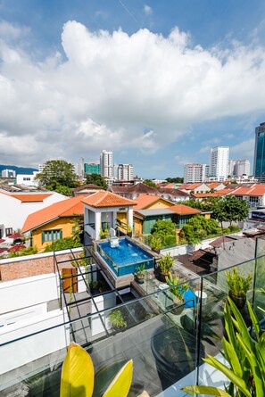 Uma piscina no terraço com guarda-corpo de vidro, rodeada por vegetação e edifícios com telhados vermelhos.