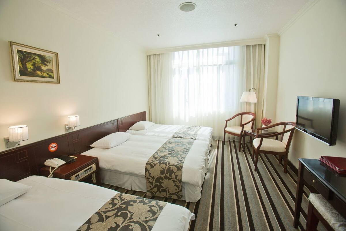Quarto de hotel com duas camas, uma mesa de trabalho com telefone, uma cadeira, televisão e janela com cortinas.