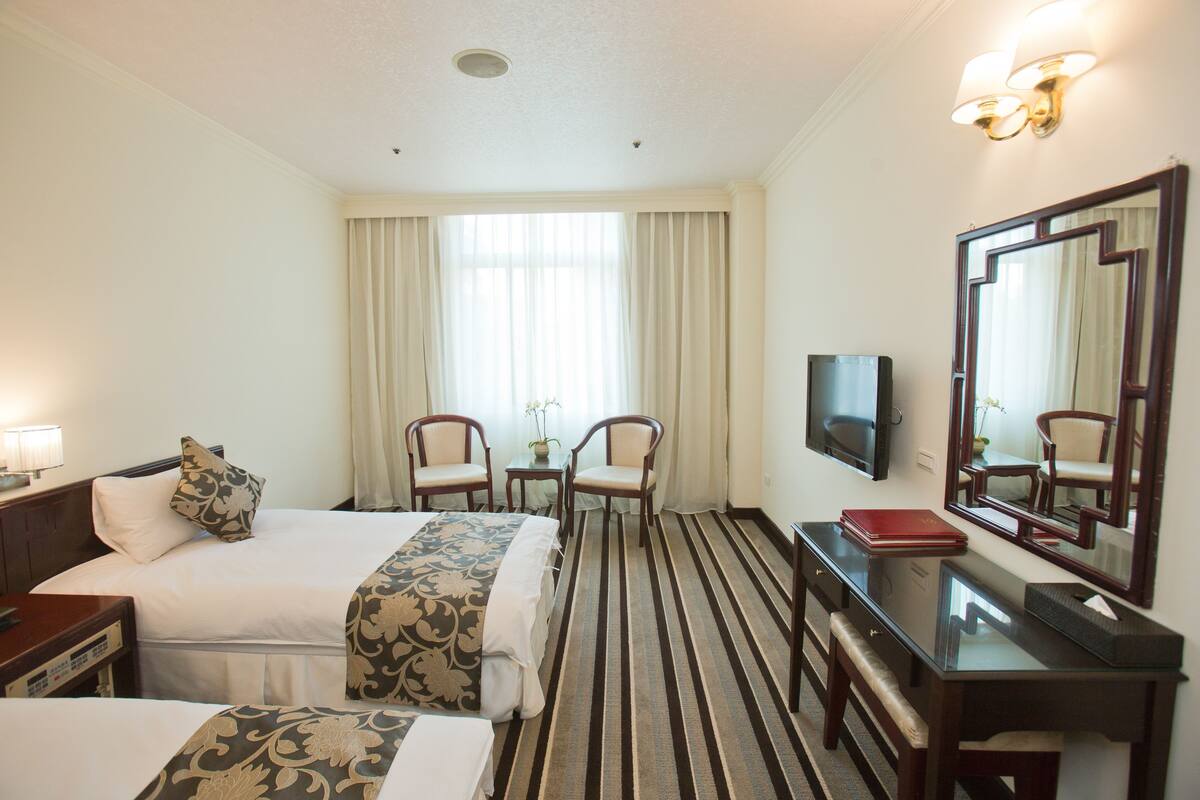 Quarto de hotel com duas camas, uma escrivaninha, uma cadeira, uma televisão e um espelho grande.