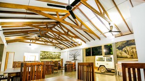 Uma sala de jantar ampla com vigas de madeira e ventiladores de teto, um SUV branco estacionado junto à janela, e obras de arte nas paredes.