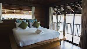 Uma cama grande com roupa de cama branca e almofadas verdes, um banco de madeira e vista para uma varanda com cortinas.