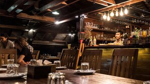 Interior de restaurante com mesas e cadeiras de madeira, área de bar com diversos garrafas e copos, e uma pessoa trabalhando atrás do balcão.