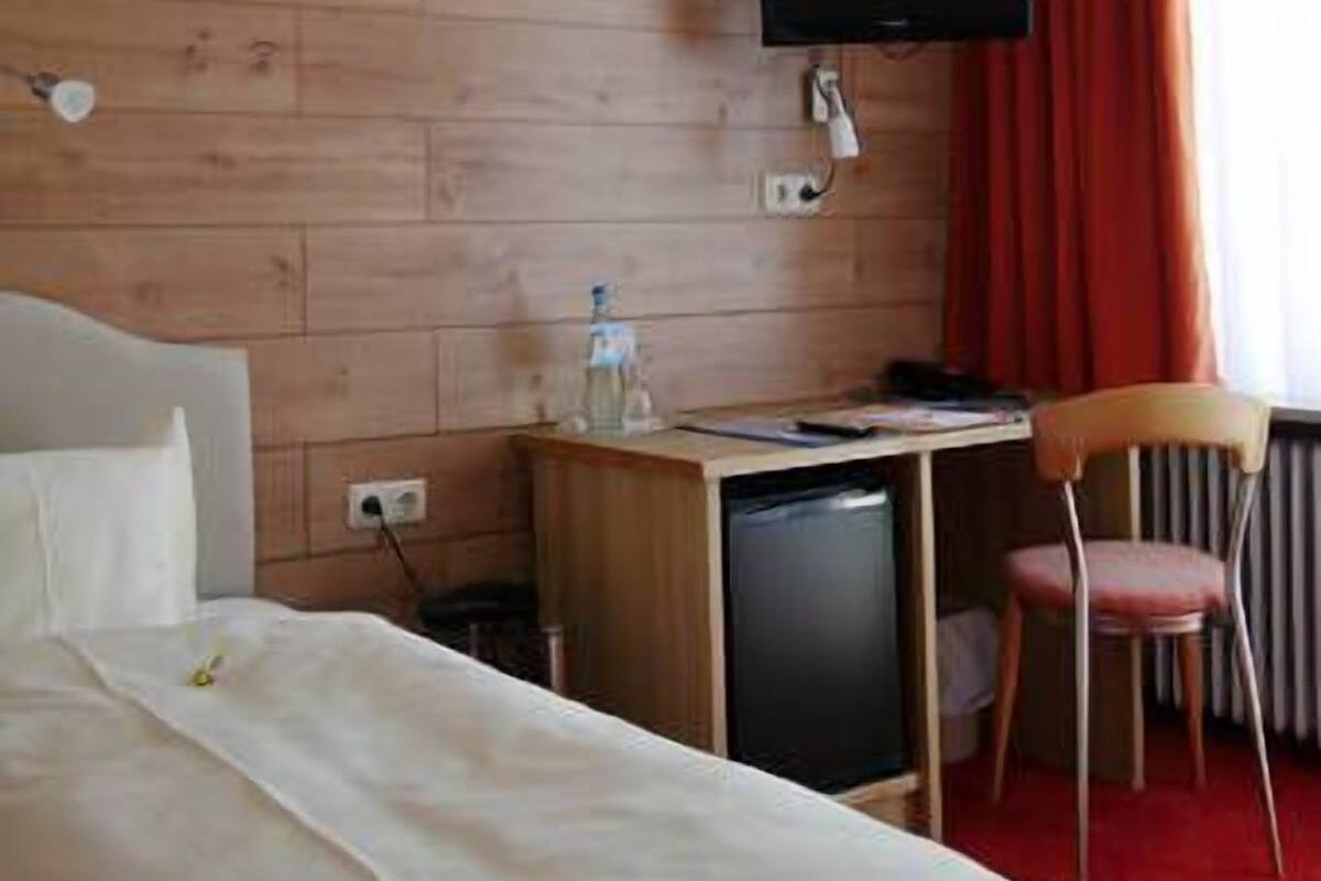 Quarto de hotel com parede de painel de madeira, cama com roupa de cama branca, escrivaninha com cadeira, televisão montada acima da escrivaninha e janela com cortinas laranja.