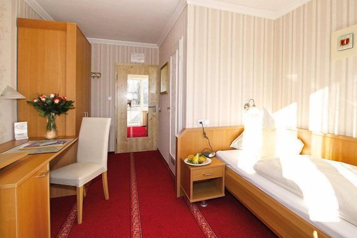 Quarto de hotel com cama, escrivaninha, cadeira, mesa de cabeceira com abajur e uma pequena mesa com frutas.