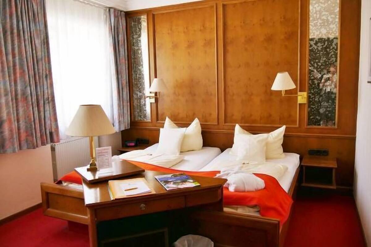 Quarto de hotel com cabeceira de madeira, duas camas com roupa de cama branca, uma escrivaninha com luminária e vista para uma janela com cortinas.
