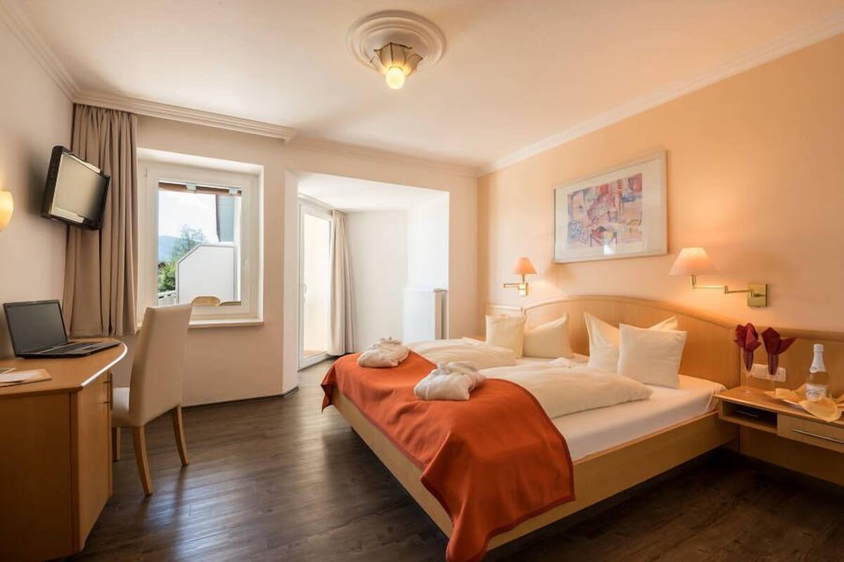 Quarto de hotel com cama, escrivaninha, cadeira, televisão e janela.