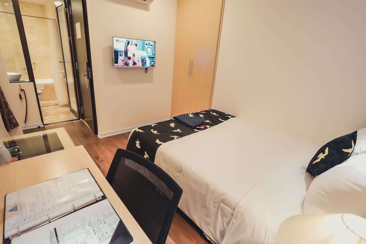 Quarto de hotel com uma cama, uma mesa com cadeira, TV na parede e banheiro com chuveiro e vaso sanitário.