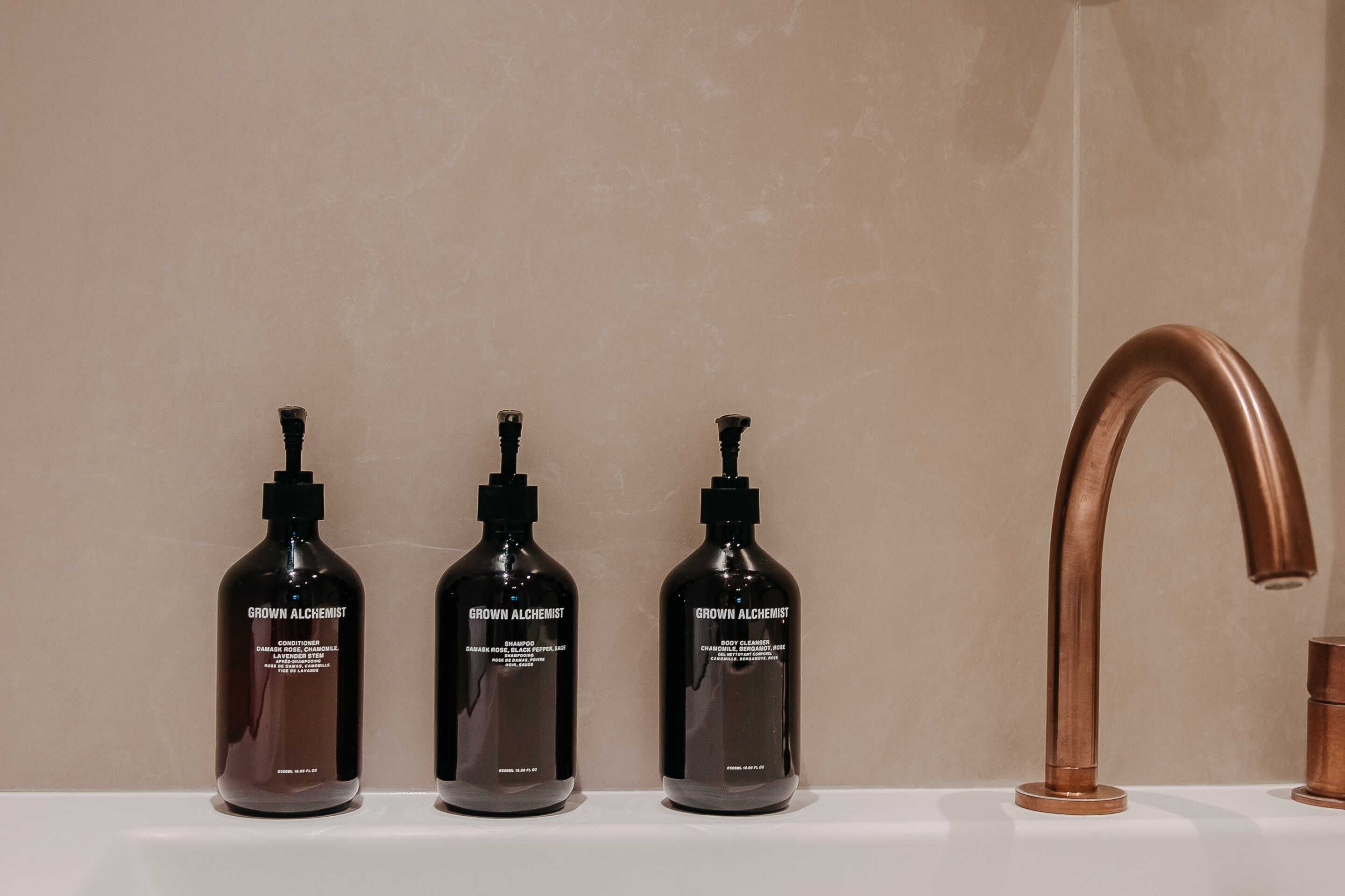 Três frascos de produtos 'Grown Alchemist' sobre uma superfície branca, ao lado de uma torneira de cobre.