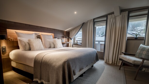 Quarto de hotel moderno com uma cama grande, uma mesinha de cabeceira, uma cadeira e vista para uma paisagem nevada pela janela.