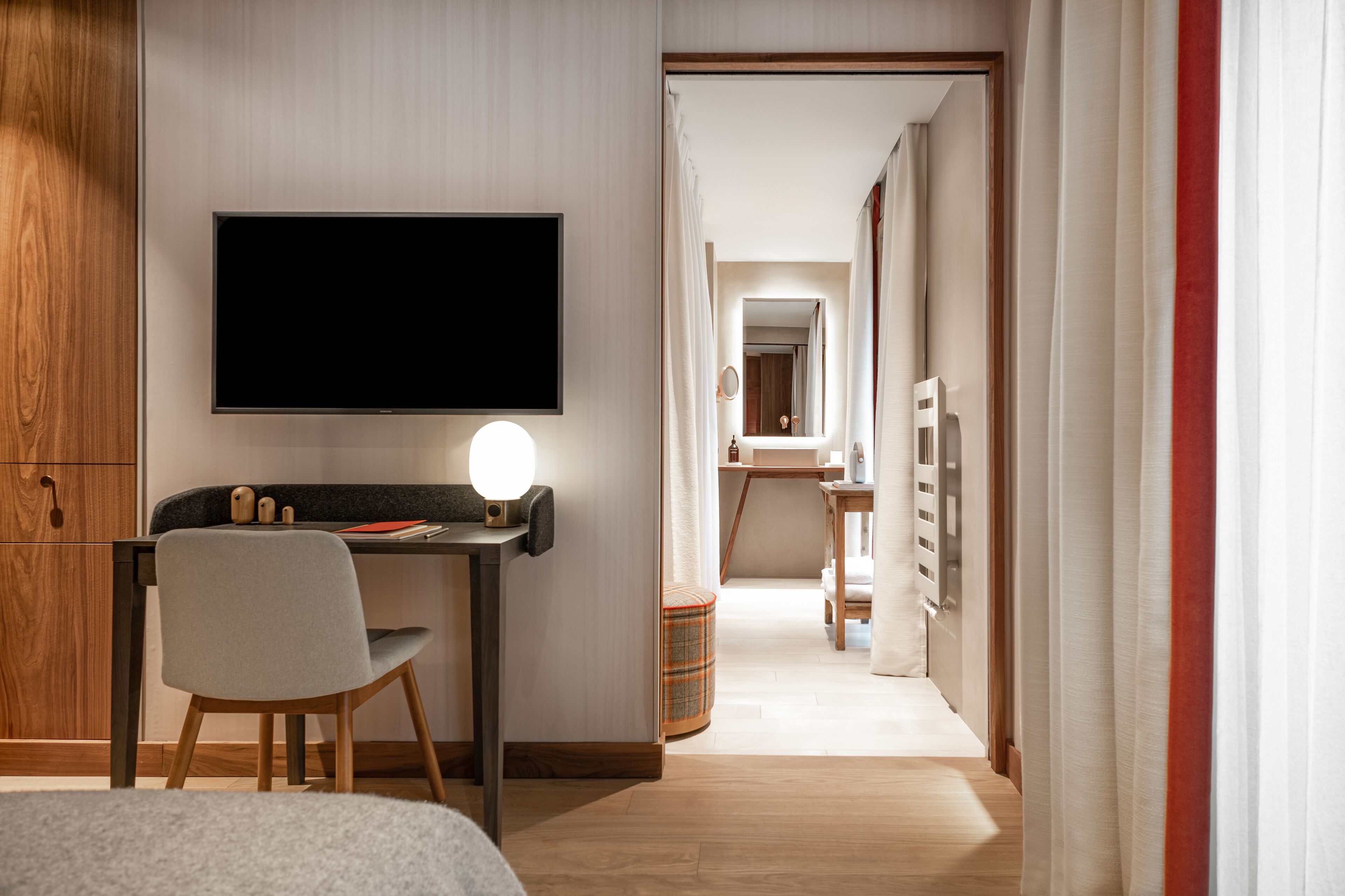 Quarto de hotel com televisão de tela plana na parede, uma escrivaninha com luminária, uma cadeira e uma cama com cabeceira branca.