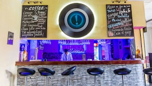 Uma área de bar com uma luminária circular central, um quadro negro com menu de café e pizzas, e um bartender no balcão.