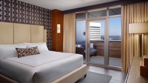 Quarto de hotel com uma cama grande, vista através de uma porta de correr para uma varanda ao ar livre, e uma cabeceira de madeira.