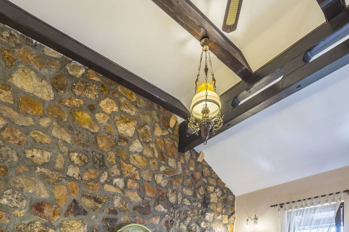 Quarto com parede de pedra, vigas de madeira, cama com roupa de cama floral, mesa de cabeceira com abajur e cômoda com espelho.
