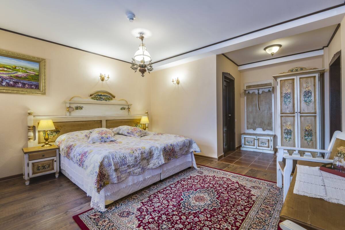 Quarto com uma cama grande, uma mesinha de cabeceira, um abajur, um candelabro, um tapete com padrão vermelho e branco e um guarda-roupa de madeira.