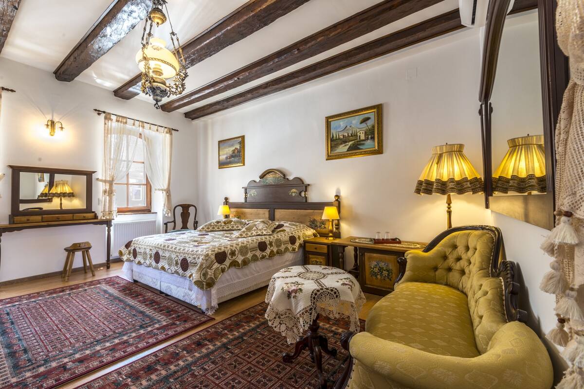 Quarto tradicional com cama, duas mesas de cabeceira, uma cadeira, uma mesinha e um espelho.