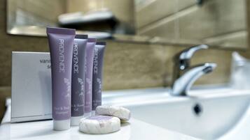 Casa de banho com lavatório branco, três frascos de shampoo e gel de banho Provence e uma barra de sabão.