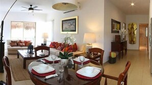 Uma sala de jantar com uma mesa redonda posta para quatro pessoas, rodeada por cadeiras de madeira. Há um ventilador de teto acima da mesa e um quadro na parede à esquerda.
