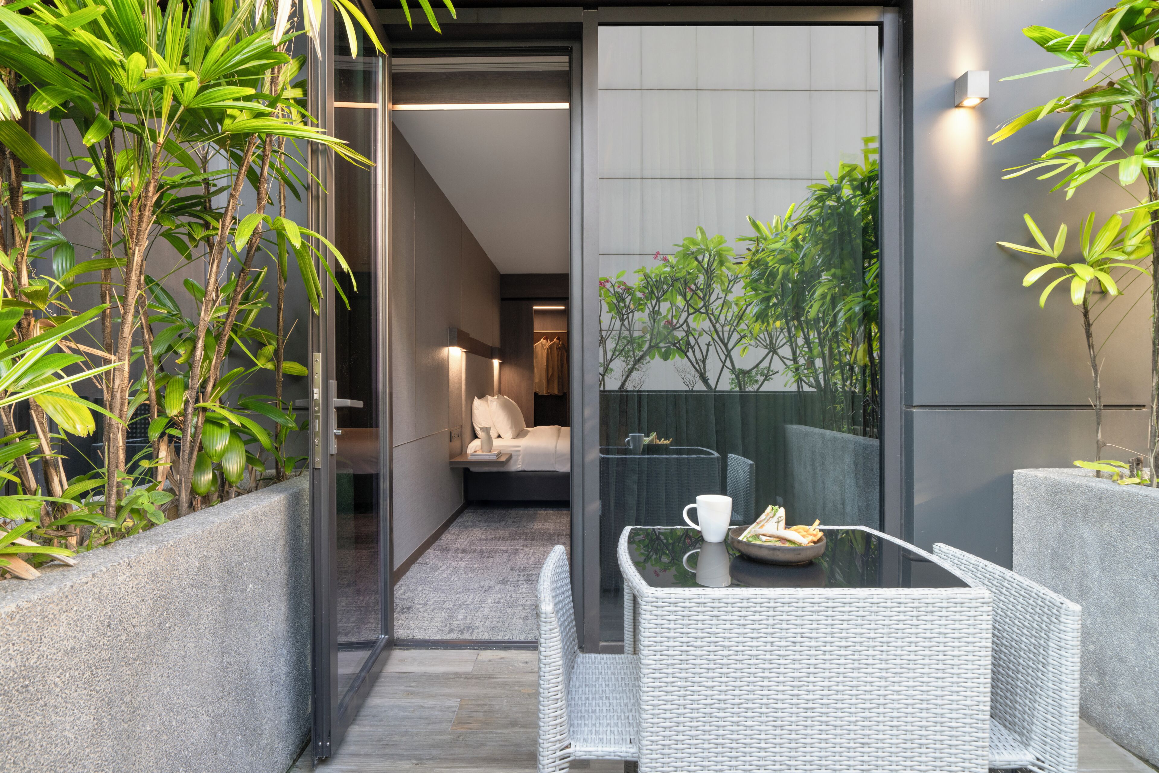Quarto de hotel moderno com divisória de vidro, área de refeições com mobiliário de vime e plantas dentro.