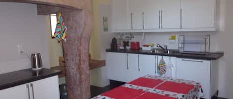Uma cozinha compacta com armários brancos, chaleira de aço inoxidável, toalha de mesa estampada em vermelho e branco, e um banquinho.