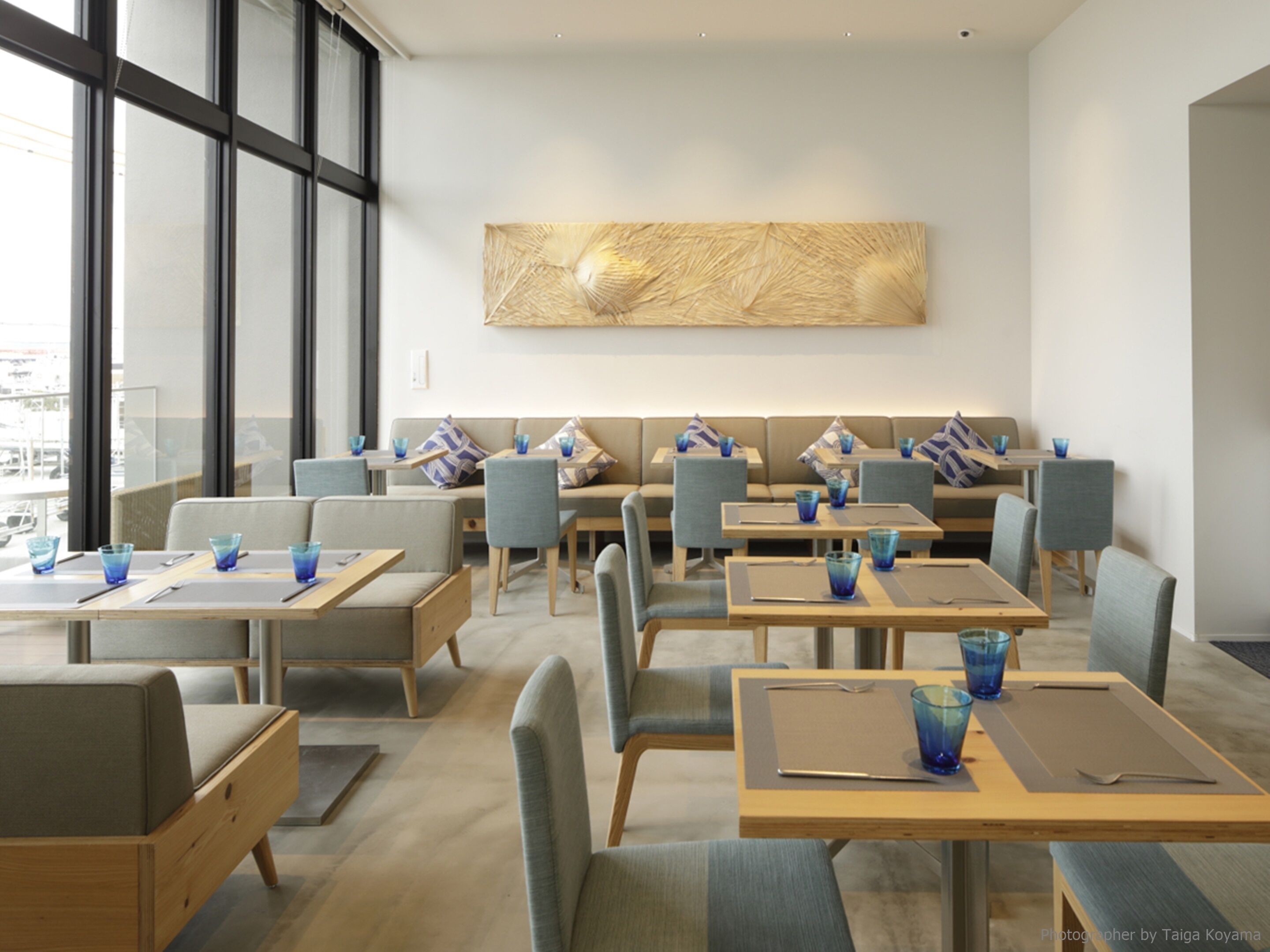 Uma sala de jantar moderna com mesas e cadeiras, um quadro abstrato grande na parede e janelas amplas.