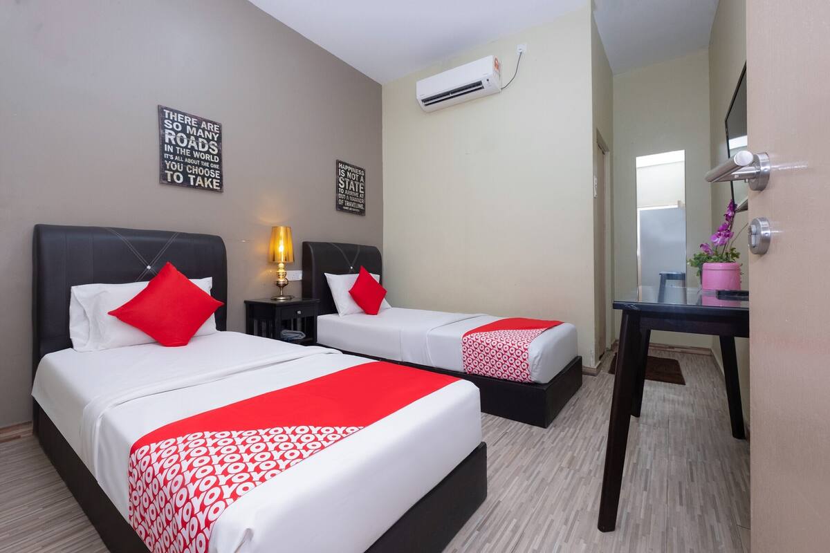 Quarto de hotel com duas camas, cada uma com roupa de cama branca e detalhes vermelhos, mesa de cabeceira com abajur, e escrivaninha com espelho e planta em vaso.