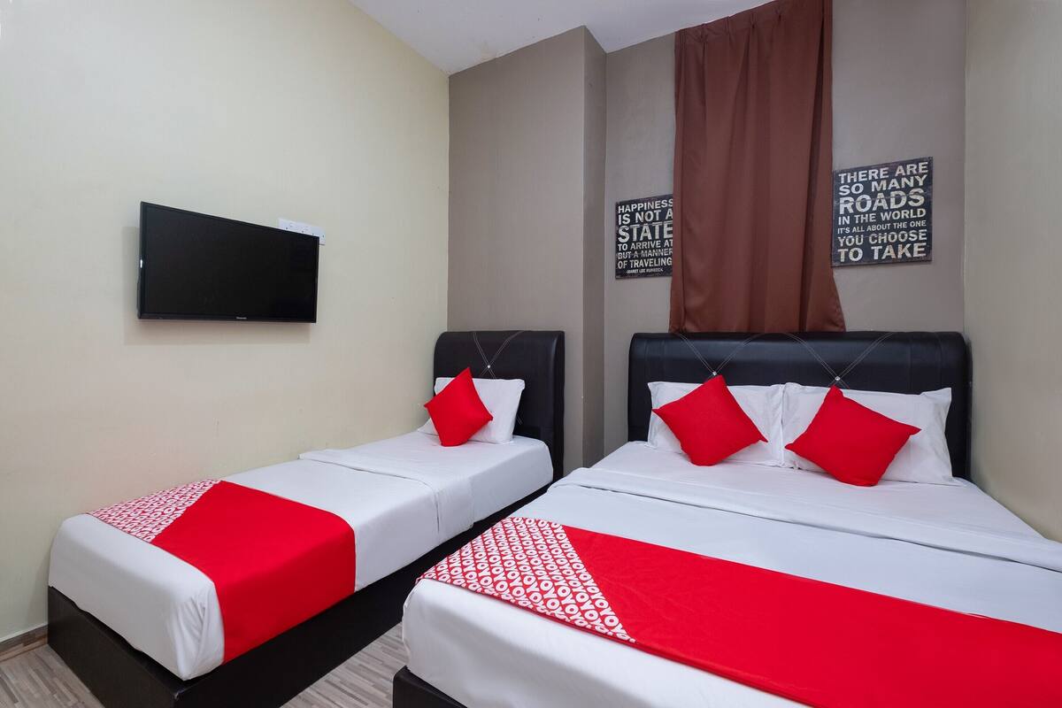 Quarto de hotel com duas camas, cada uma com roupa de cama vermelha e branca, uma TV de tela plana na parede e frases motivacionais decorando a parede.