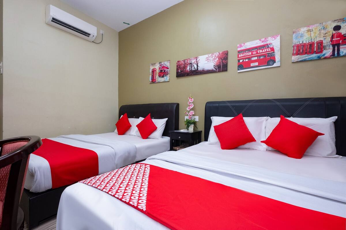 Quarto de hotel com duas camas, cada uma com roupa de cama e manta em vermelho e branco. Há um ar condicionado fixado na parede e três quadros representando um ônibus vermelho, um ônibus de dois andares vermelho e uma pessoa com uma mala.