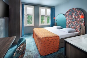 Quarto de hotel com cama, escrivaninha, cadeira, banheiro e janela com cortinas.