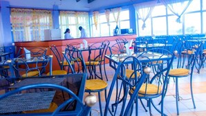 Um restaurante com cadeiras e mesas de metal azul, um balcão com eletrodoméstico branco e grandes janelas com cortinas.