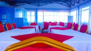 Uma sala de conferências com mesas e cadeiras dispostas em fileiras, amplas janelas com cortinas e um teto alto com vigas aparentes.