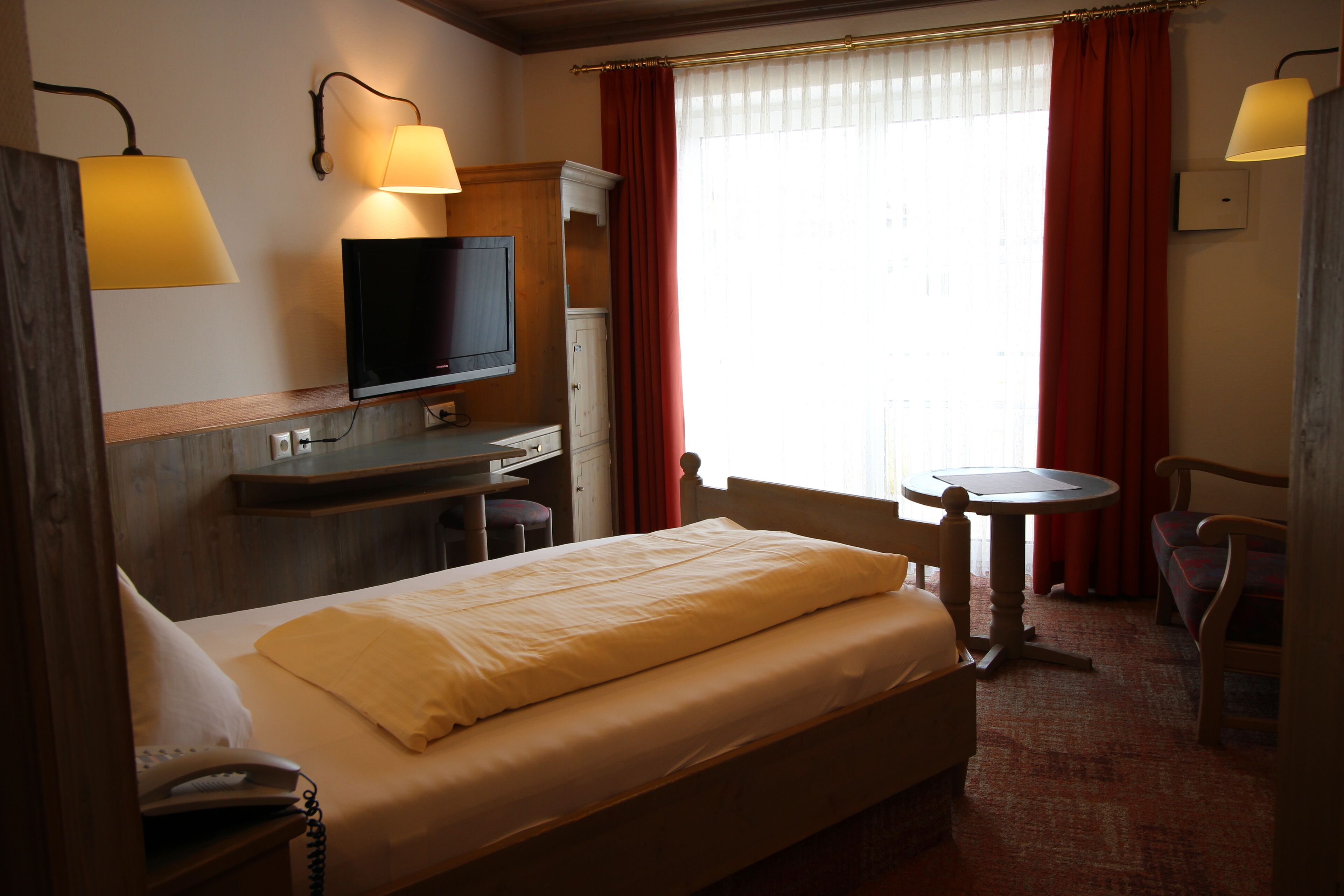 Quarto de hotel com uma cama, escrivaninha com televisão, uma cadeira, mesa pequena e janela com cortinas.