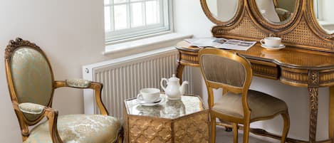 Um boudoir de estilo antigo com mobiliário ornamentado, incluindo uma toucador com três espelhos, uma cadeira e uma pequena mesa com um bule e chávena.