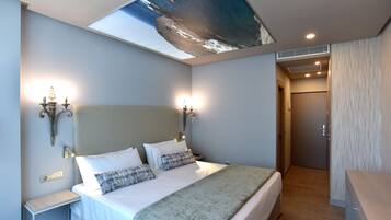 Quarto de hotel moderno com cama grande, duas mesas de cabeceira, um mural no teto e vista para o mar.
