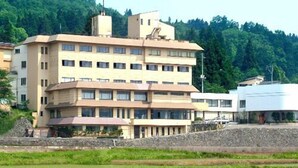 Um hotel de vários andares com telhado plano e chaminé central, rodeado por campos verdes e uma colina arborizada.