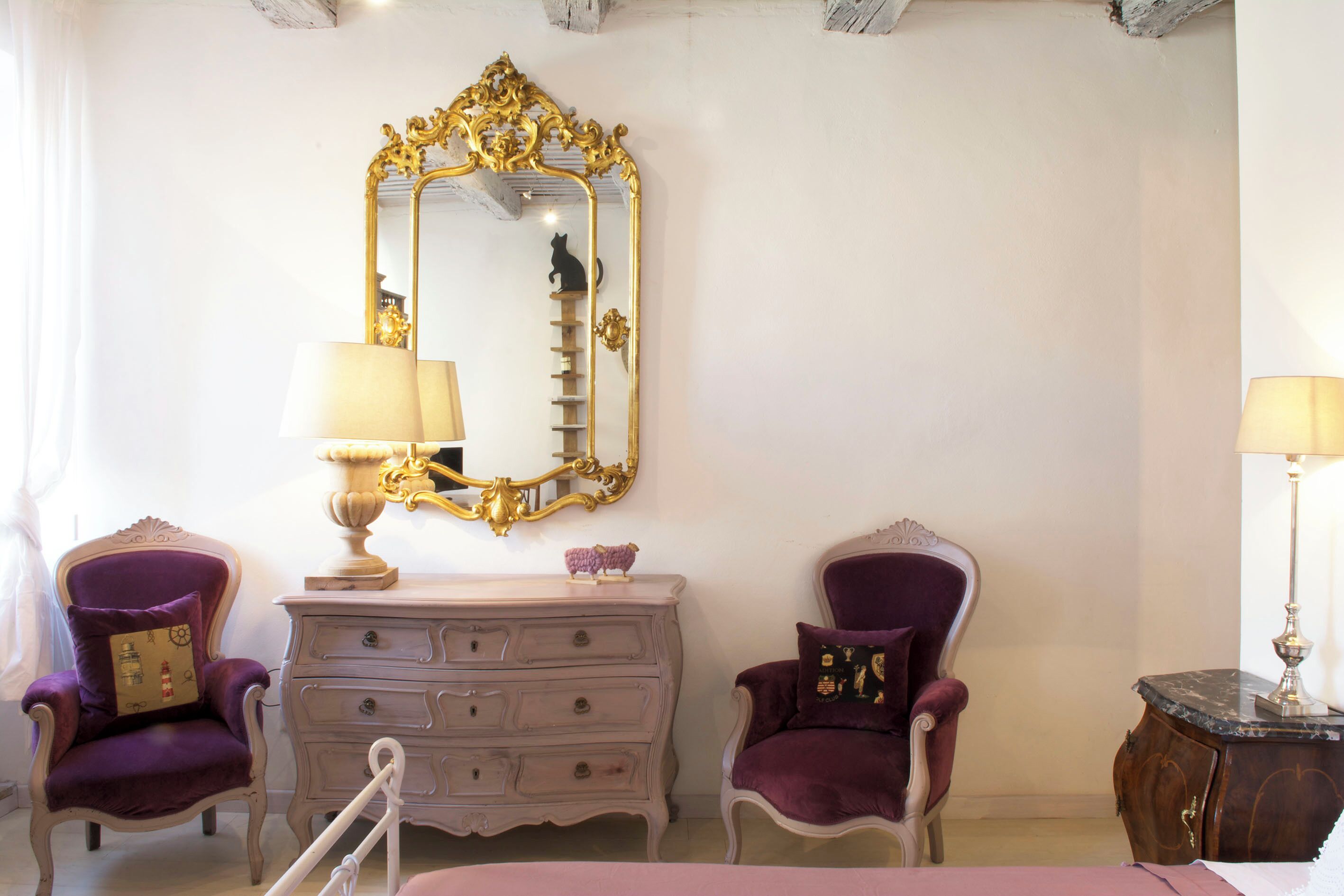 Um quarto com um espelho grande e ornamentado, duas poltronas roxas, uma cômoda branca e uma cama com colcha rosa.