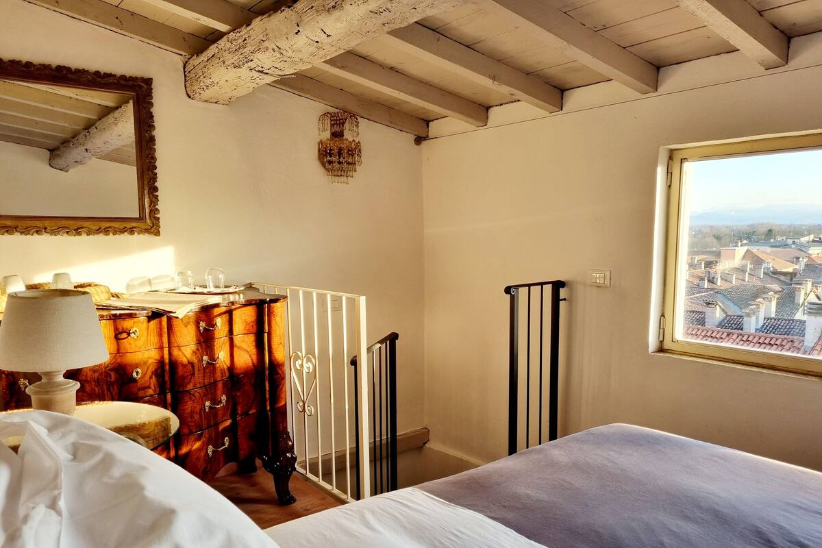 Quarto com cama, penteadeira, espelho, lustre e janela com vista para a cidade.
