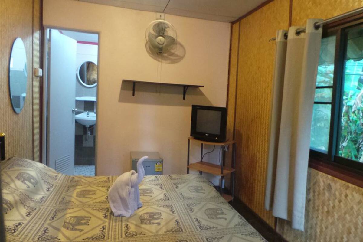 Quarto de hotel com cama, TV sobre suporte, ventoinha, janela com cortinas e porta para o banheiro.