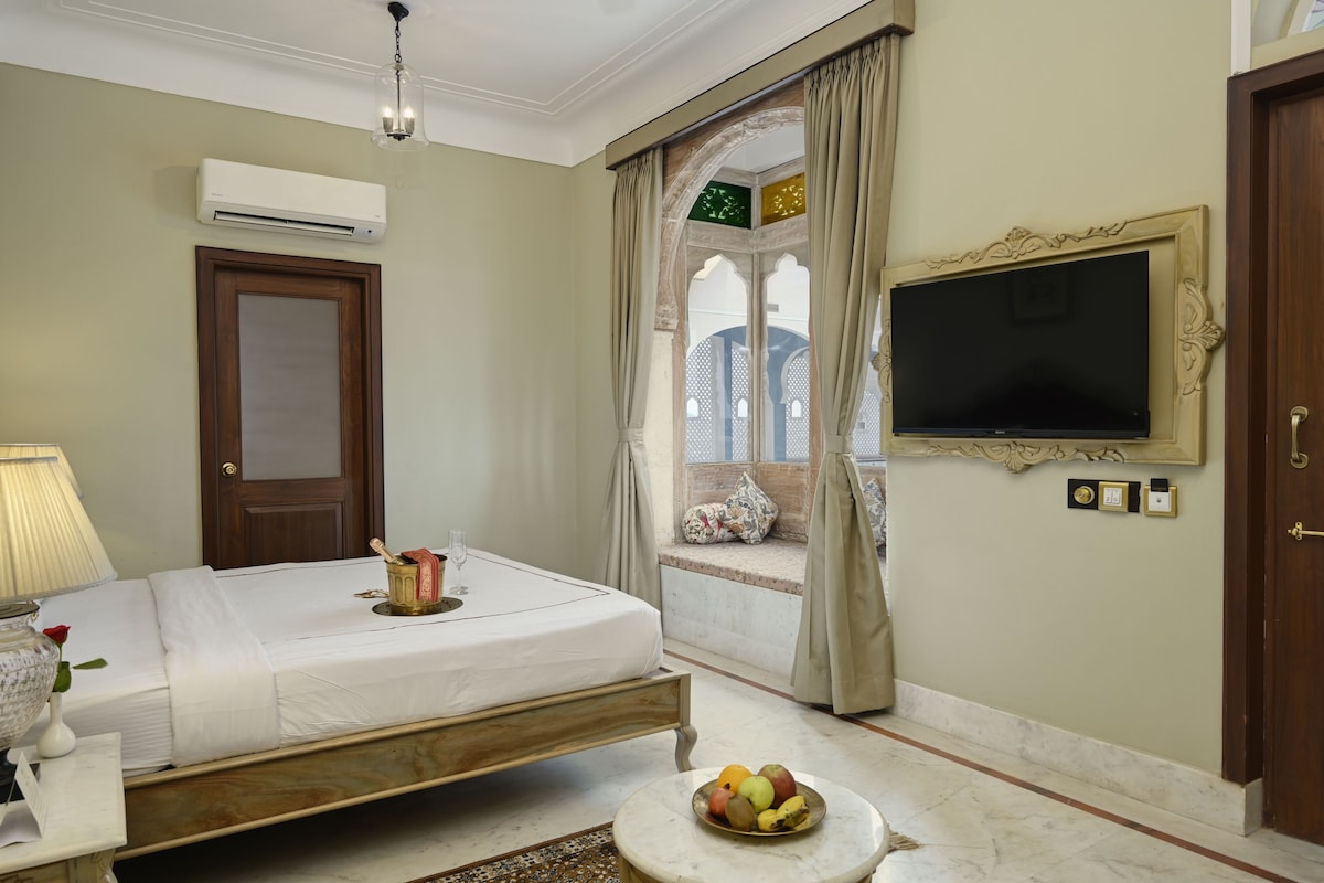 Quarto com cama, televisão, ar condicionado, janela com cortinas e uma pequena mesa com frutas.