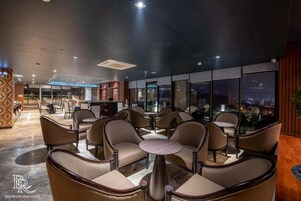 Um hall de hotel moderno com poltronas de couro, área de bar e grandes janelas com vista para a cidade.
