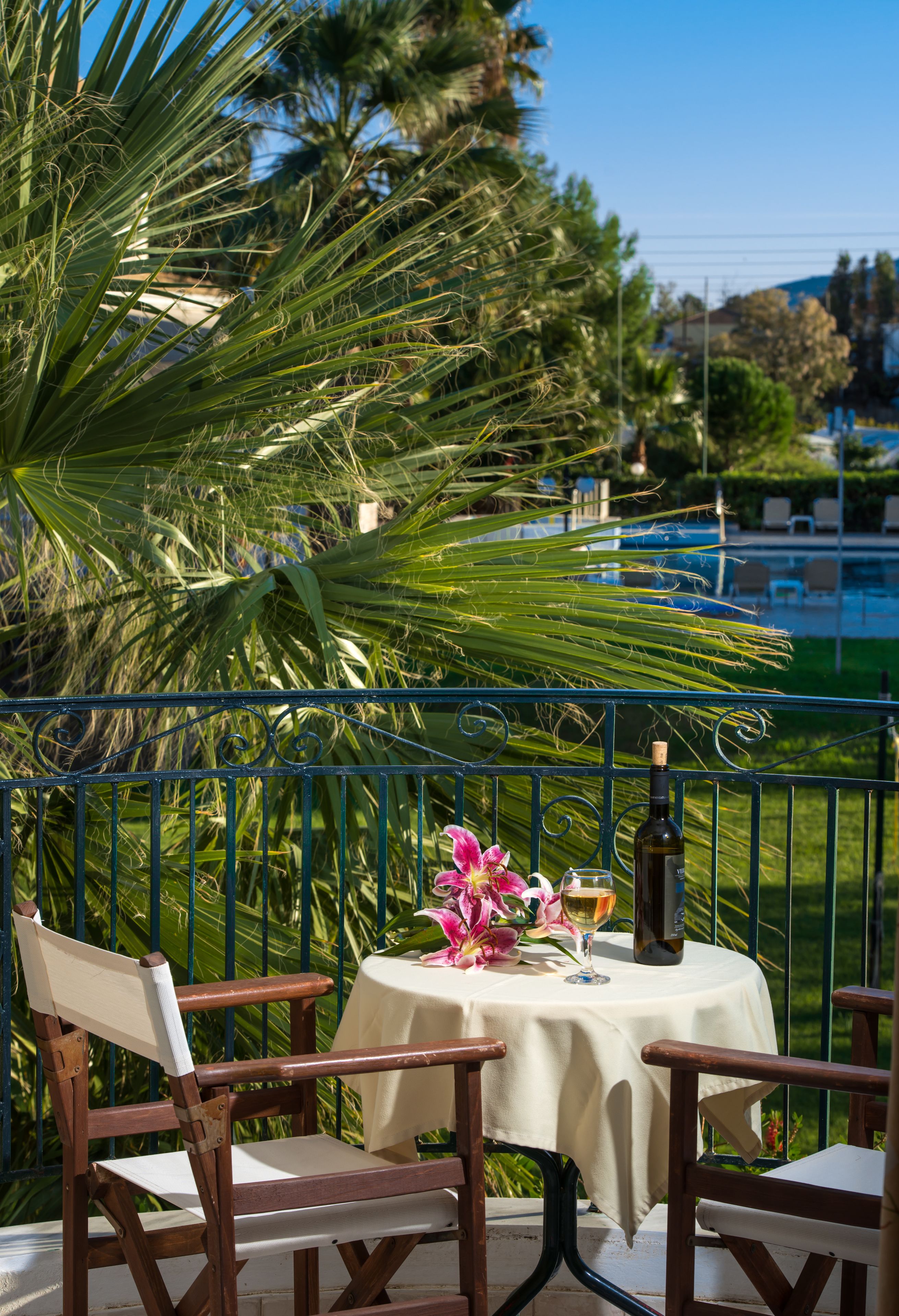 Uma mesa com uma garrafa e um copo, rodeada de cadeiras, com vista para uma piscina e palmeiras.