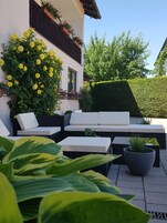Uma área externa com assentos, incluindo um sofá e vasos de plantas, um arbusto e um edifício com caixas de flores.