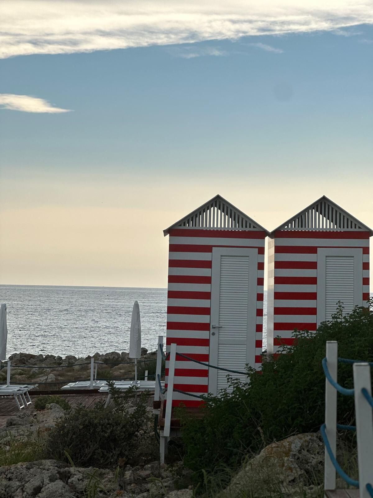 Duas cabanas de praia com paredes listradas em vermelho e branco e janelas com persianas brancas, localizadas perto do mar com uma linha costeira rochosa ao primeiro plano.