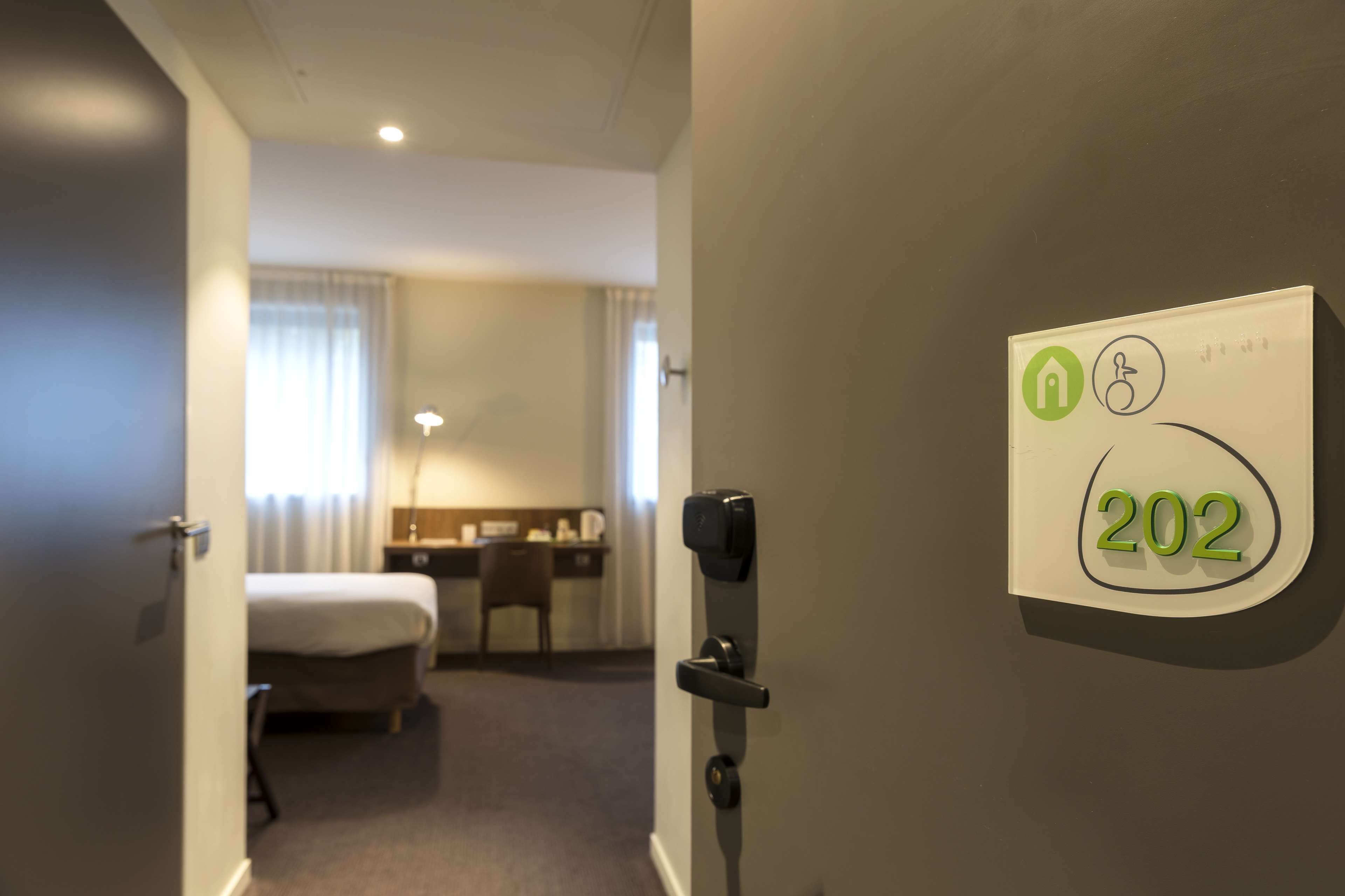 Quarto de hotel com cama, escrivaninha e porta com o número indicado.