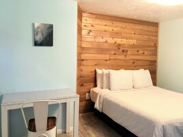 Quarto com cabeceira de madeira, cama com roupa de cama branca, uma mesinha com cadeira e um quadro pendurado na parede.