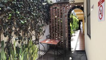 Um corredor externo estreito com parede de tijolos coberta de hera, estrutura de madeira treliçada e uma mesa com cadeiras.