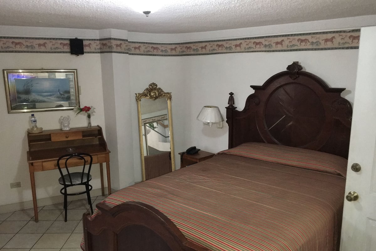 Quarto com cama de madeira, escrivaninha com cadeira, espelho e quadro na parede.