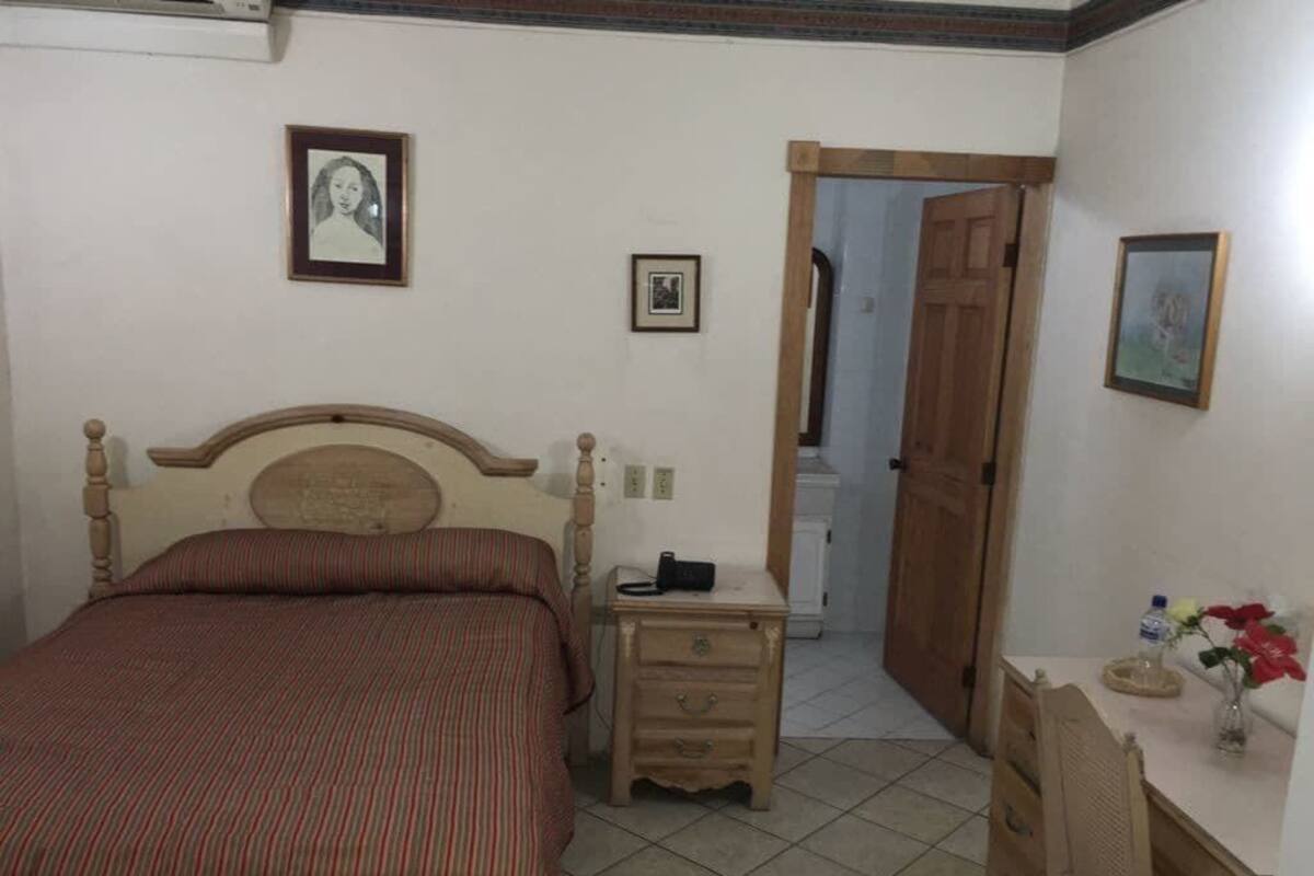 Um quarto com cama, mesa de cabeceira, cômoda e porta que dá para outro espaço.