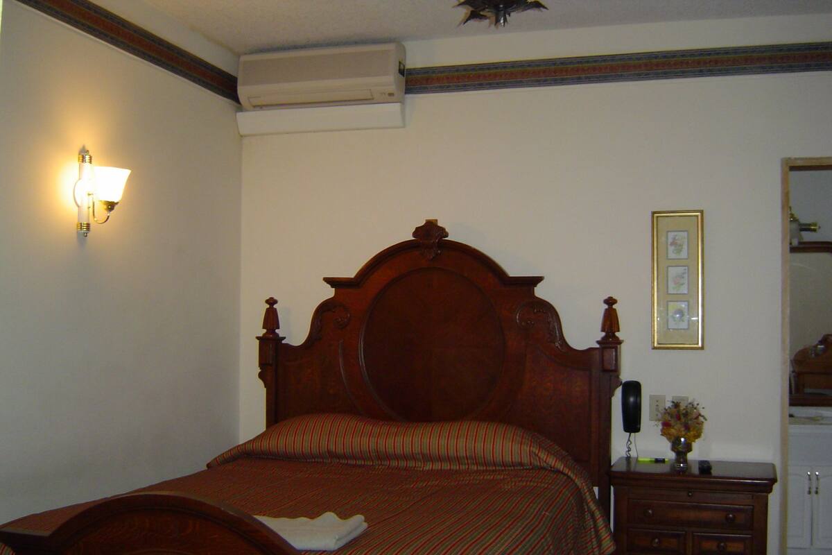 Uma cama de madeira com cabeceira, uma mesa de cabeceira com um vaso e um candeeiro fixo na parede.