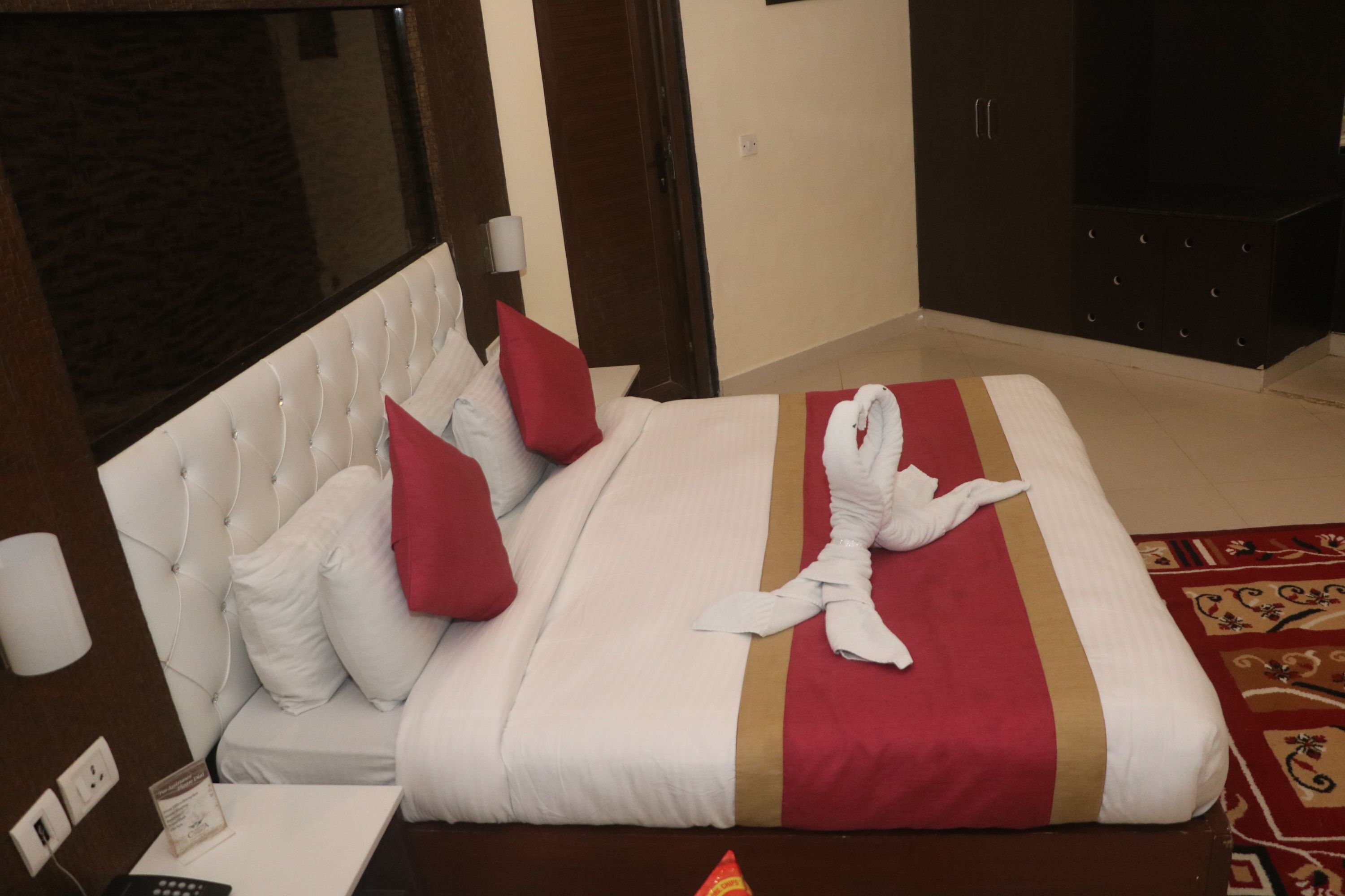 Quarto de hotel com uma cama grande, roupa de cama branca, almofadas vermelhas, edredom vermelho e dourado, toalha branca sobre a cama, mesa de cabeceira com controle remoto e luminária fixada na parede.